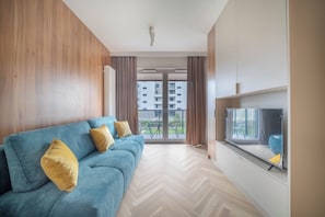 Uma sala de estar moderna com um sofá azul, televisão e vista para um prédio residencial.