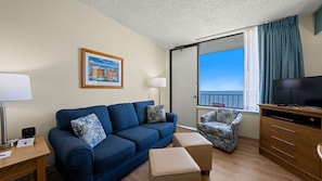 Quarto com sofá azul, poltrona estampada, televisão e janela com vista.
