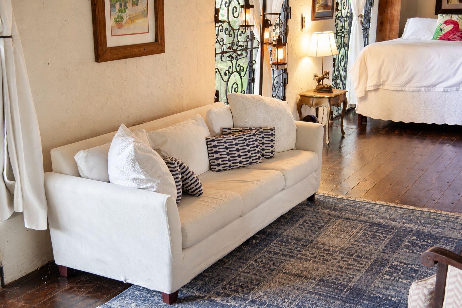 Quarto com sofá branco, almofadas estampadas, tapete azul estampado, piso de madeira e cama com cobertura branca.