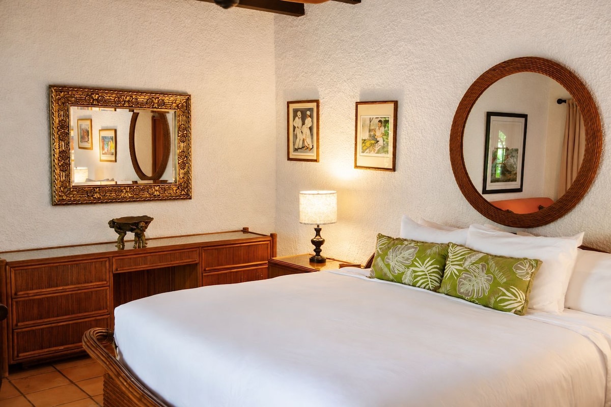 Quarto de hotel com cama grande, penteadeira de madeira, espelho e quadros nas paredes.