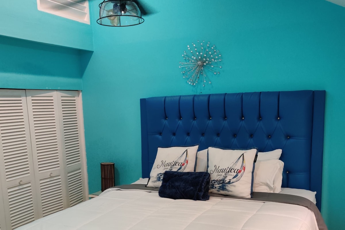 Quarto com cabeceira azul, roupa de cama branca e ventilador de teto decorativo.