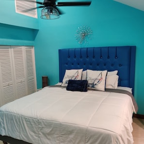 Quarto com cabeceira azul, roupa de cama branca e ventilador de teto decorativo.