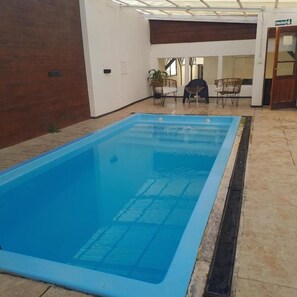 Piscina interior retangular com água azul cristalina, rodeada por um chão ladrilhado e uma parede de madeira.