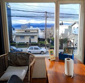 Quarto com vista para uma rua residencial, uma cadeira de vime, uma mesa de madeira com um rolo de papel higiénico e um quadro emoldurado sobre a mesa.