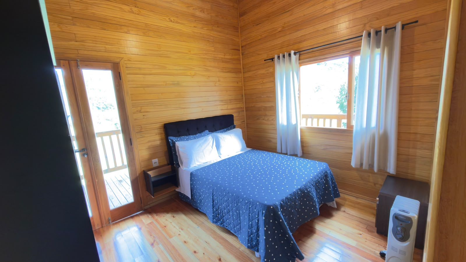 Quarto com painéis de madeira, uma cama, uma mesinha de cabeceira e uma janela com cortinas.