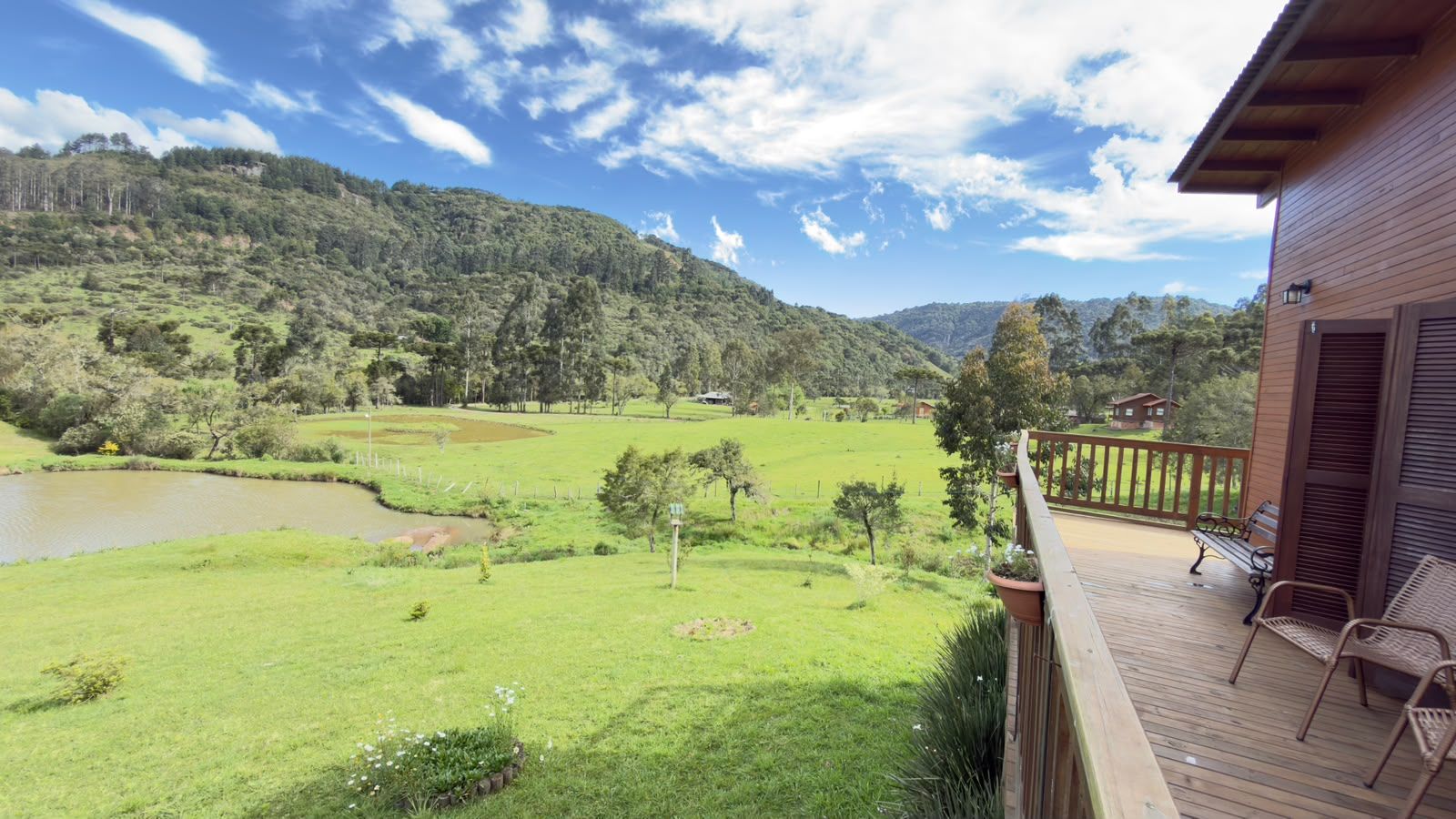 Um deck de madeira com móveis para área externa, com vista para uma área gramada com lago e árvores, e colinas ao fundo.