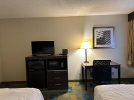 Quarto de hotel com cama, escrivaninha com abajur, televisão e micro-ondas.