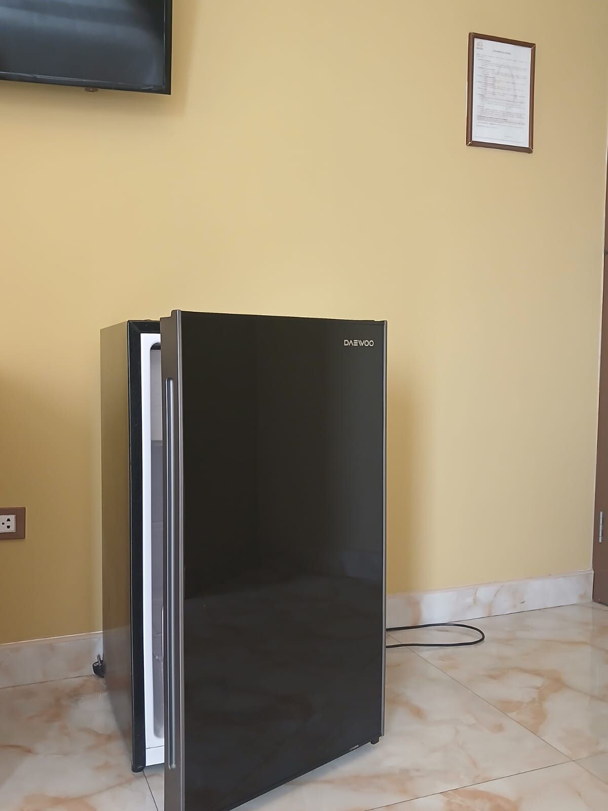 Uma geladeira preta com painel de controle na porta, uma televisão fixada na parede e um certificado emoldurado.
