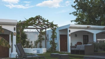 Área de pátio com pérgola, mobiliário externo e um edifício com fachada branca e portas em arco.
