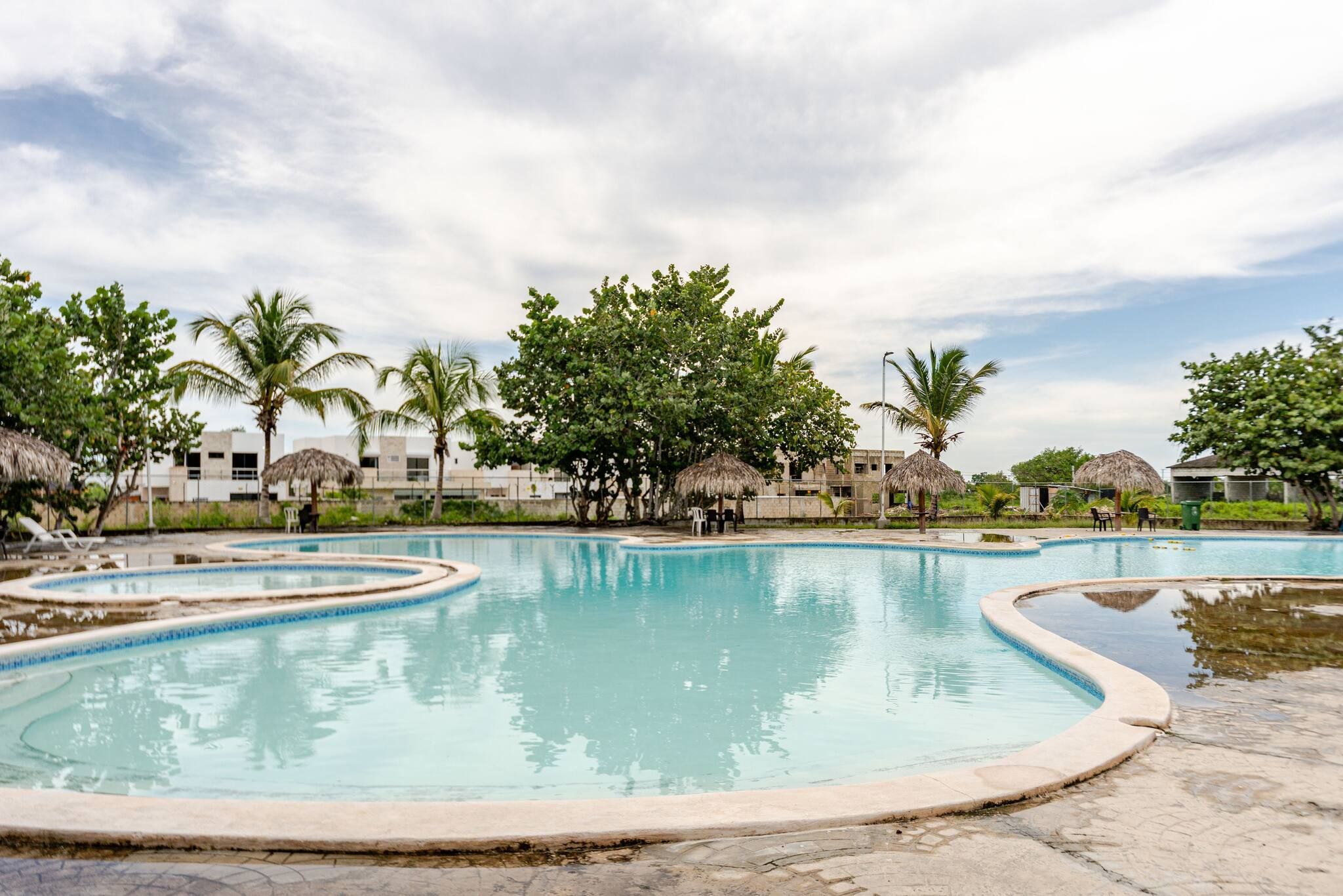 Piscina com várias áreas, rodeada por palmeiras e construções com telhados de palha.