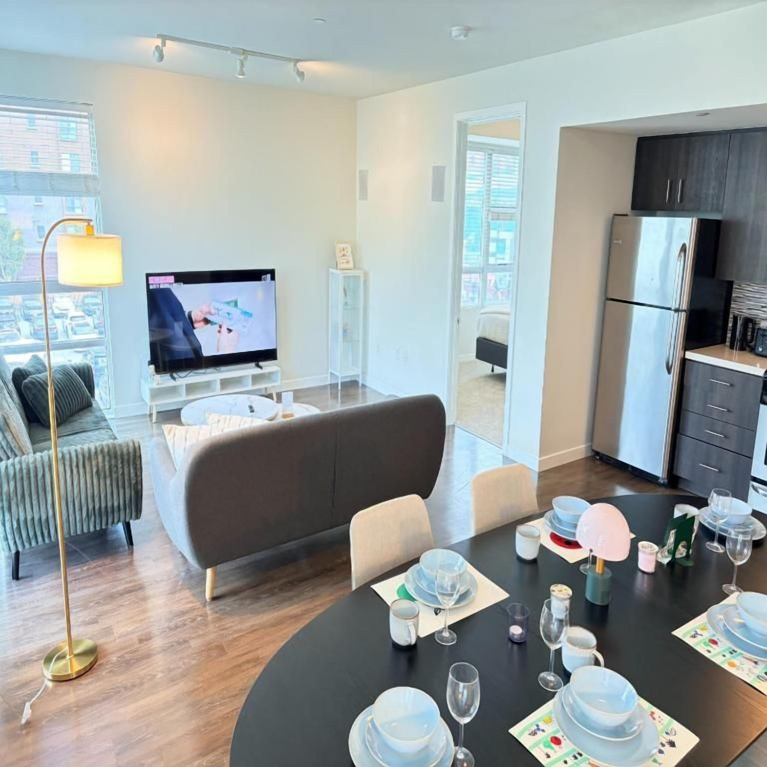 Uma sala de estar moderna com sofá, mesa de jantar posta para quatro pessoas, televisão e área de cozinha com geladeira.