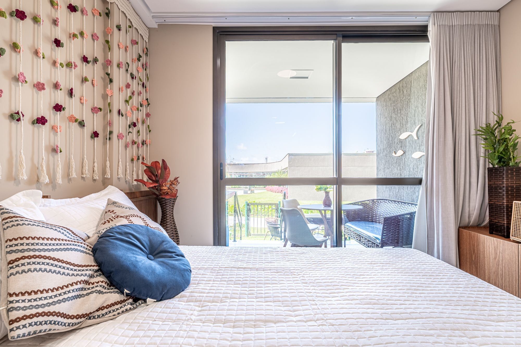 Quarto com cama grande, vista para o exterior por uma porta de correr e um painel de parede decorativo em macramé.