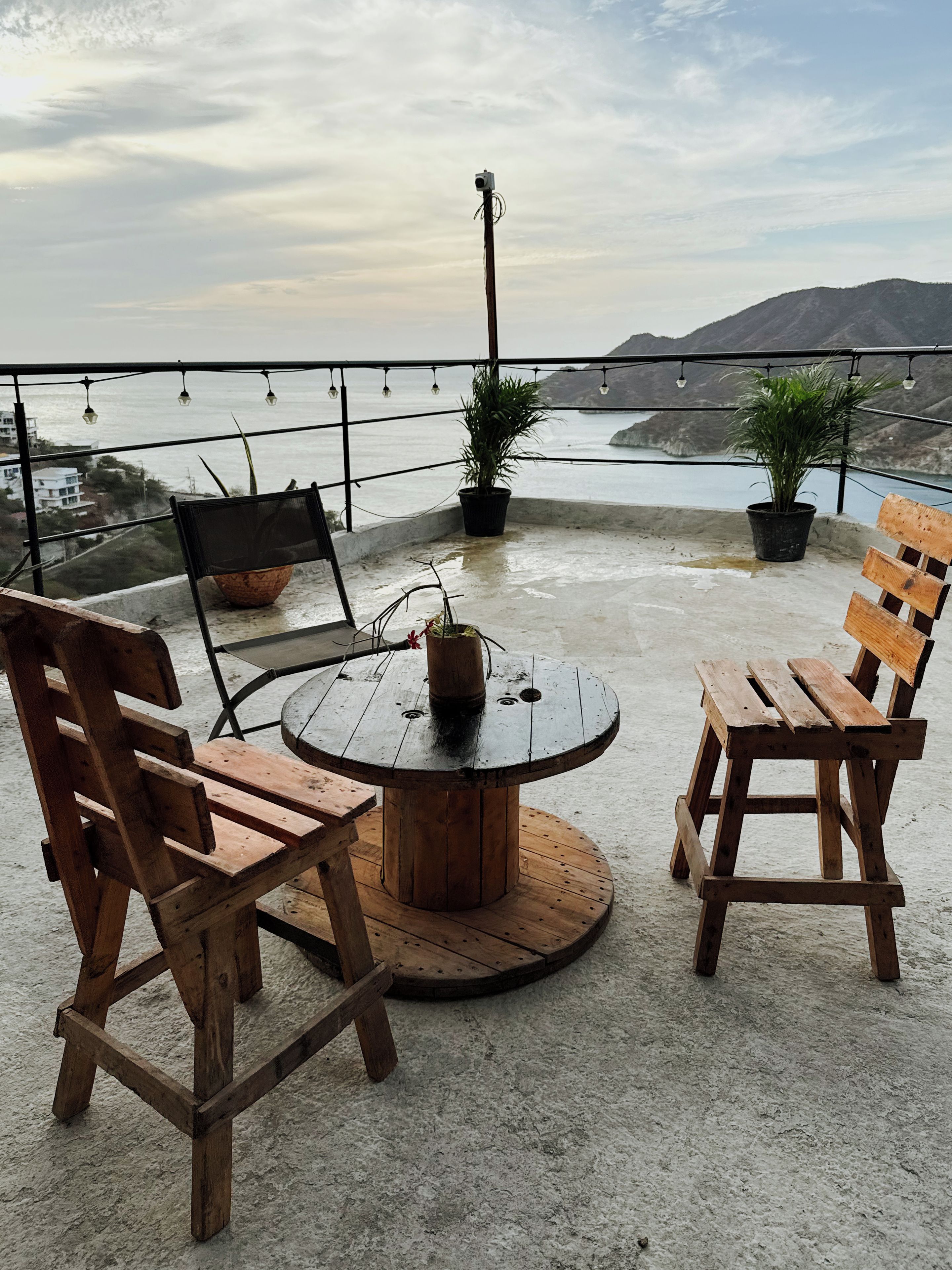 Mesa e cadeiras de madeira numa varanda com vista para um corpo de água e montanhas ao longe.