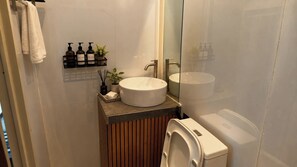 Casa de banho com toucador em madeira, lavatório branco, espelho e sanita. Há artigos de higiene pessoal no toucador e uma toalha pendurada na parede.