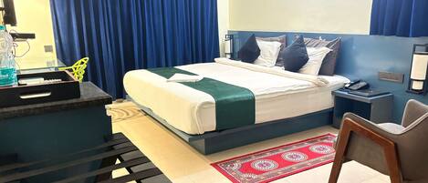 Quarto de hotel com cama, mesas de cabeceira, escrivaninha com cadeira, tapete estampado em vermelho e janela com cortinas.