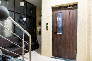 Entrada de hotel com porta de madeira, elevador de vidro e escada com corrimãos de metal.