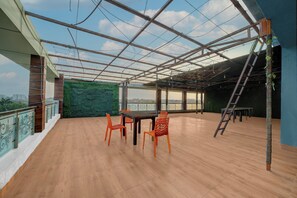 Espaço aberto com piso de madeira, mesa com cadeiras, parede verde e vista da cidade.