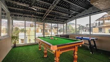 Uma mesa de bilhar com pano verde, rodeada por um espaço amplo com teto alto e janelas grandes.