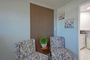 Quarto com duas poltronas estampadas, parede de ripas de madeira e uma pequena mesa com uma planta verde.