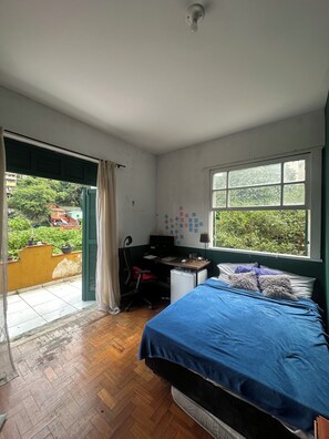 Quarto com cama, escrivaninha, cadeira, mini-frigorífico e janela com vista para área verde.