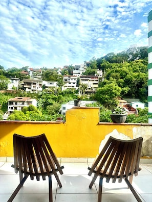 Duas cadeiras de madeira numa varanda com uma parede amarela, com vista para uma encosta com edifícios e vegetação.