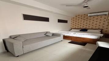 Quarto com sofá cinza, cama com roupa de cama branca, ventoinha no teto e aparelho de ar condicionado.