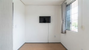 Quarto com uma televisão preta fixada na parede, uma janela com cortinas e piso de madeira.