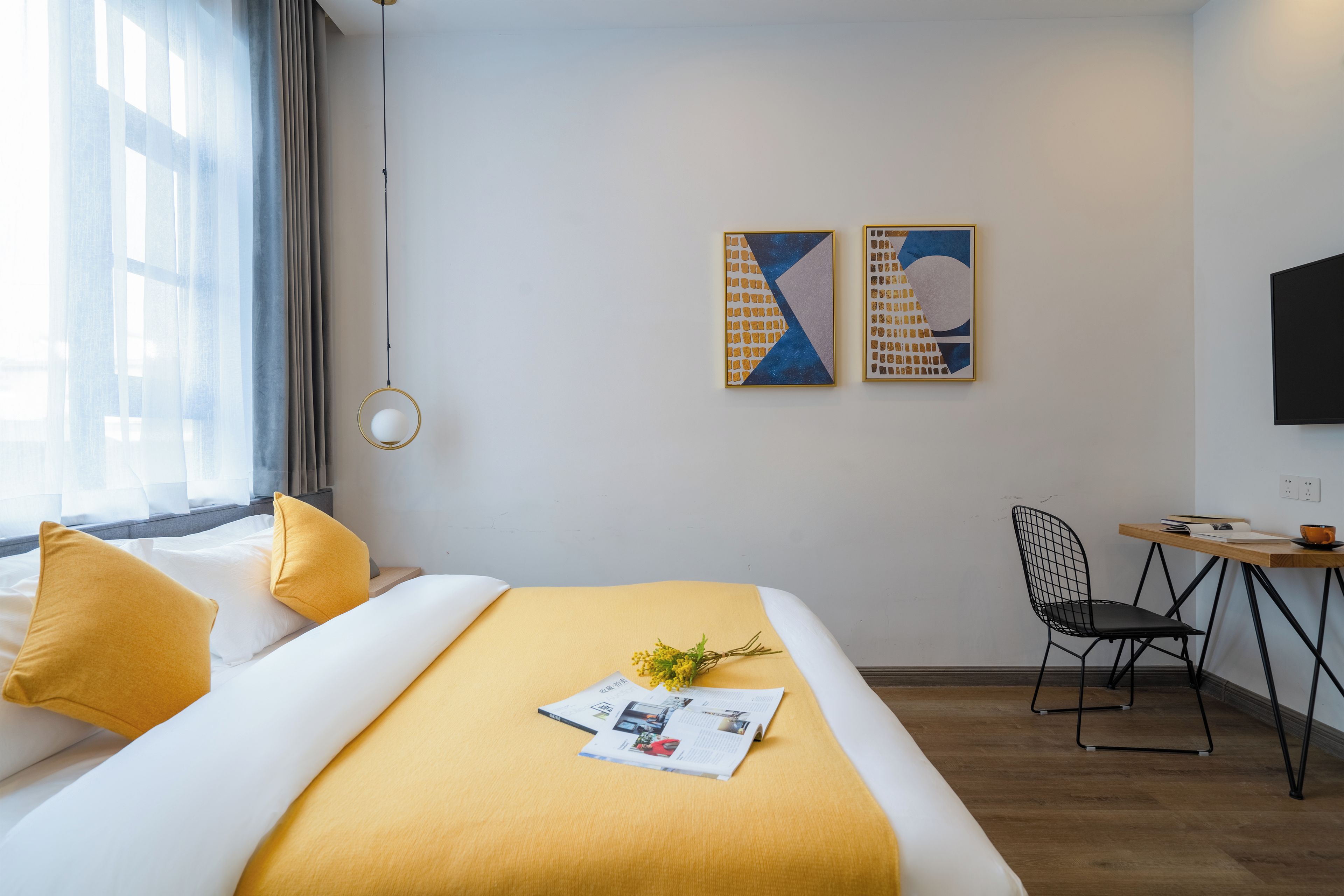 Quarto de hotel com cama, escrivaninha e duas pinturas na parede.