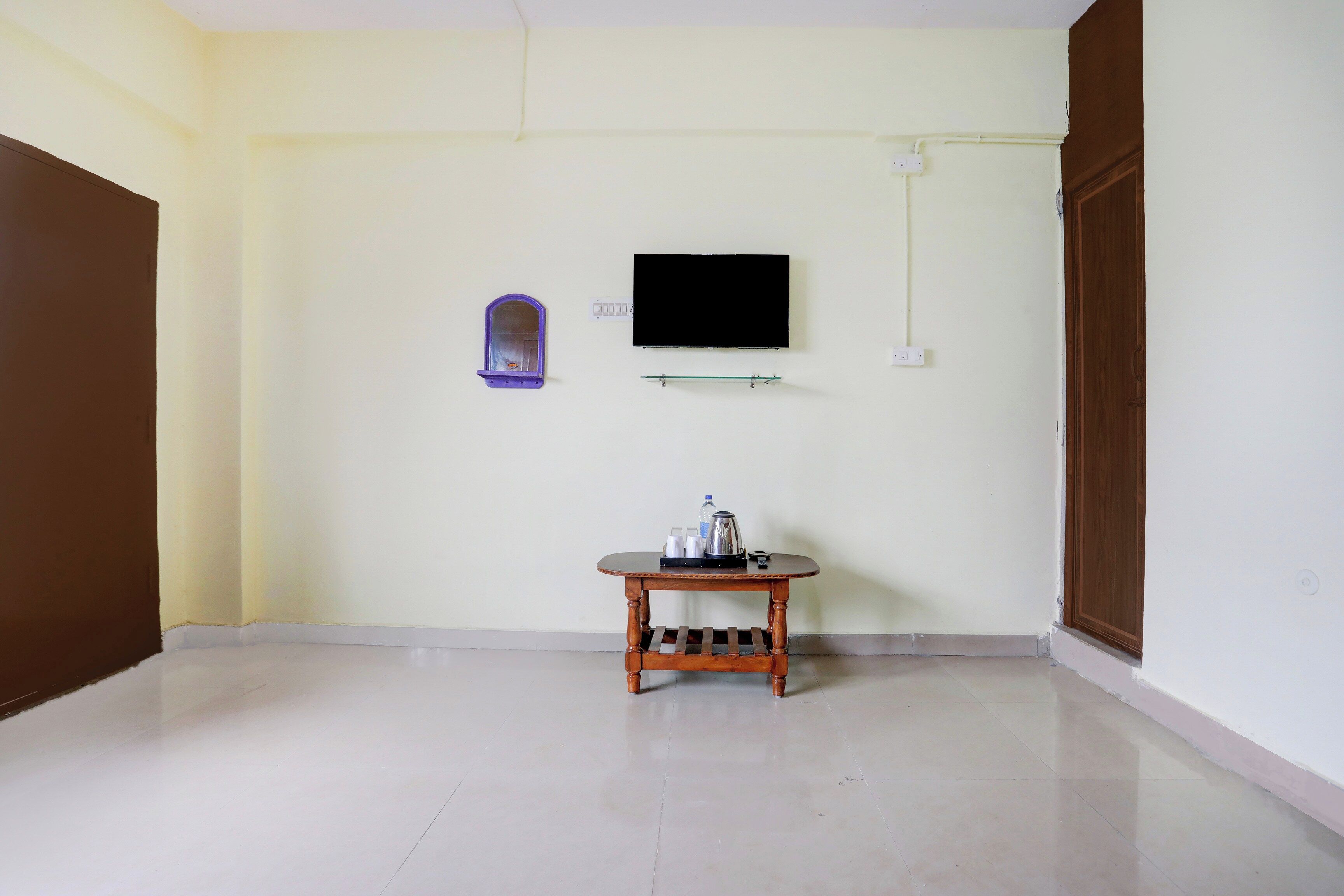 Quarto com televisão de tela plana na parede, uma mesinha com chaleira e xícaras, e uma porta à direita.