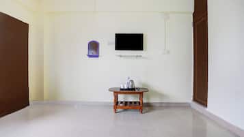 Quarto com televisão de tela plana na parede, uma mesinha com chaleira e xícaras, e uma porta à direita.