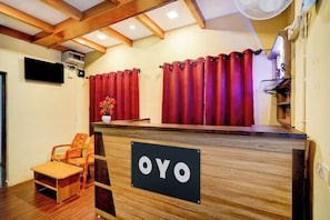 Bancada de recepção com placa indicando 'OYO' em um saguão de hotel com piso de madeira, cortinas vermelhas e ventilador de teto.
