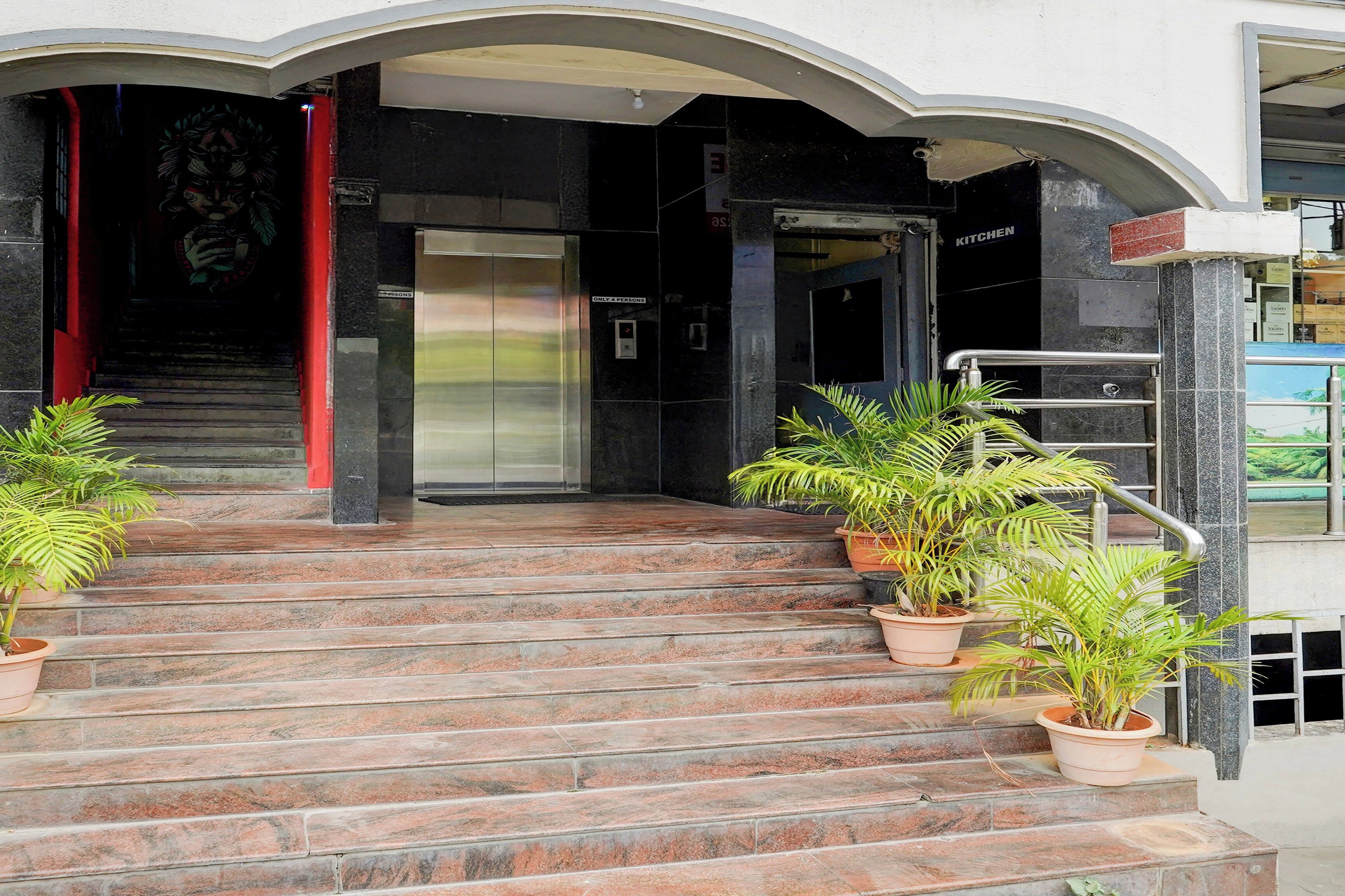 Entrada de um edifício com escadaria que leva a um elevador, plantas em vasos e uma placa indicando 'Apenas 4 pessoas'.