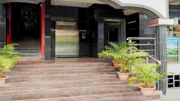 Entrada de um edifício com escadaria que leva a um elevador, plantas em vasos e uma placa indicando 'Apenas 4 pessoas'.