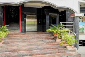 Entrada de um edifício com escadaria que leva a um elevador, plantas em vasos e uma placa indicando 'Apenas 4 pessoas'.