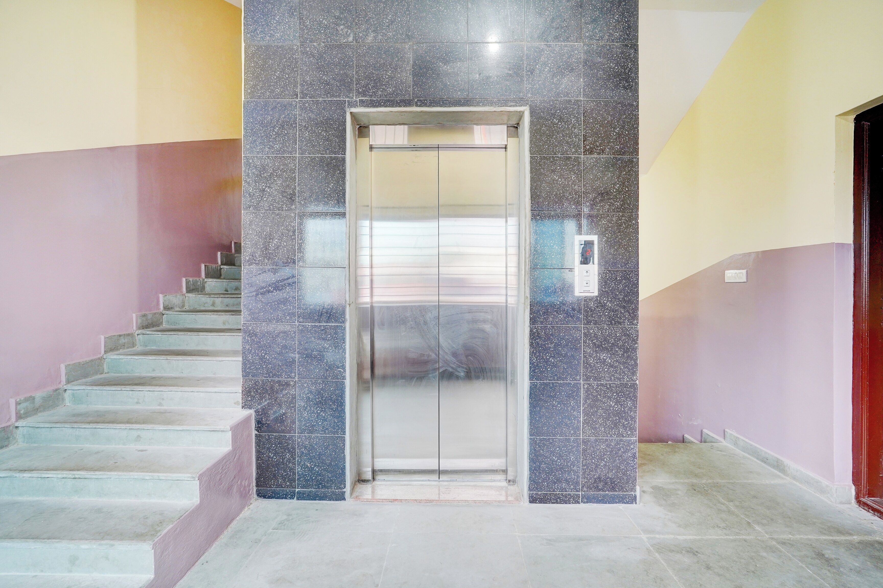 Um elevador moderno com painel de controle, próximo a uma escada com uma parede rosa de um lado e uma parede amarela do outro.