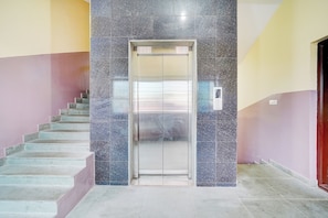 Um elevador moderno com painel de controle, próximo a uma escada com uma parede rosa de um lado e uma parede amarela do outro.