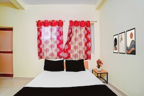 Quarto com cama, duas almofadas pretas, mesa de cabeceira com vaso de flores e janela com cortinas vermelhas.