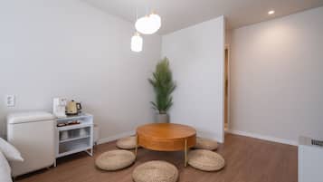 Uma sala de estar moderna com uma mesa de centro redonda de madeira, pufes trançados, um sofá branco, um armário branco com uma cafeteira e uma planta em vaso.