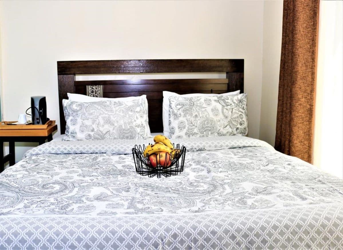 Uma cama bem arrumada com roupa de cama estampada, uma cesta de frutas sobre a cama e uma mesinha de cabeceira de madeira com um alto-falante ao lado.