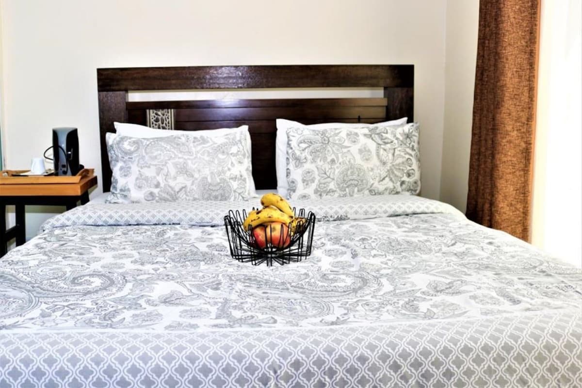 Uma cama bem arrumada com roupa de cama estampada, uma cesta de frutas sobre a cama e uma mesinha de cabeceira de madeira com um alto-falante ao lado.