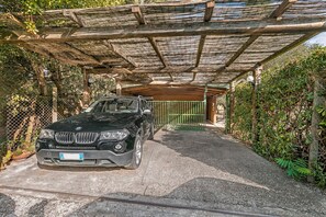 Carro preto estacionado sob uma pérgola de madeira, com um portão verde ao fundo.