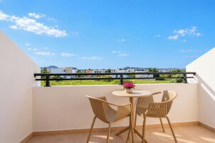 Uma varanda com mesa e cadeiras, um vaso de flores e vista para uma paisagem verde e edifícios ao longe.