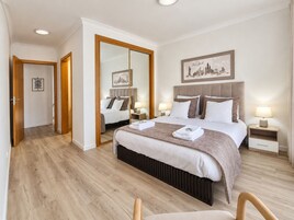 Quarto de hotel com uma cama grande, mesas de cabeceira com luminárias, um espelho e uma imagem emoldurada da paisagem urbana na parede.