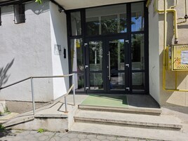 Entrada de um prédio com porta de vidro, corrimão de metal e caixa de utilidades amarela.