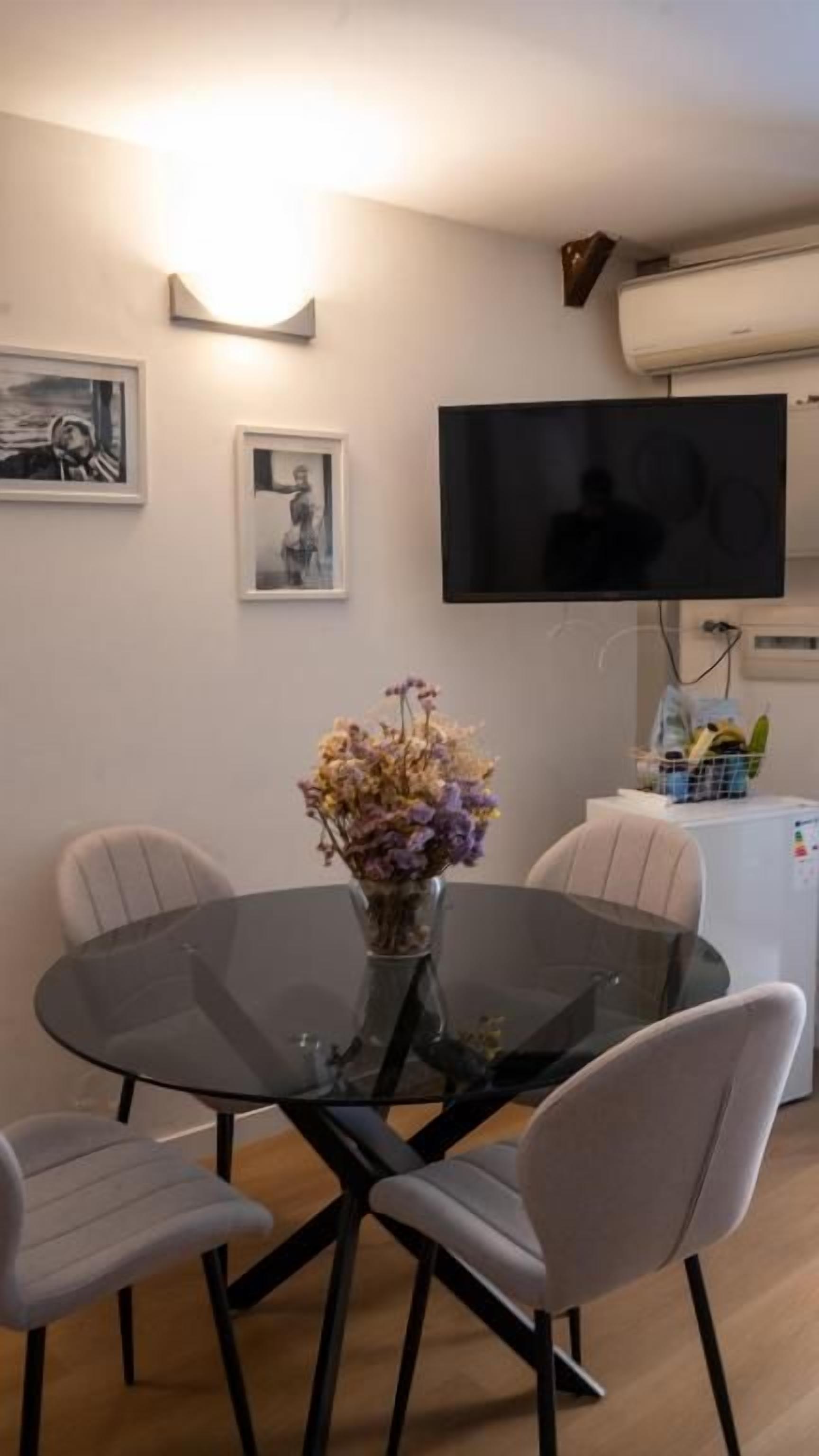 Uma área de refeições com uma mesa redonda de vidro, quatro cadeiras, um vaso com flores, uma TV de tela plana na parede e uma geladeira pequena.
