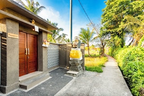 Entrada da villa com porta de madeira, placa indicativa, pequeno jardim e caminho de acesso.