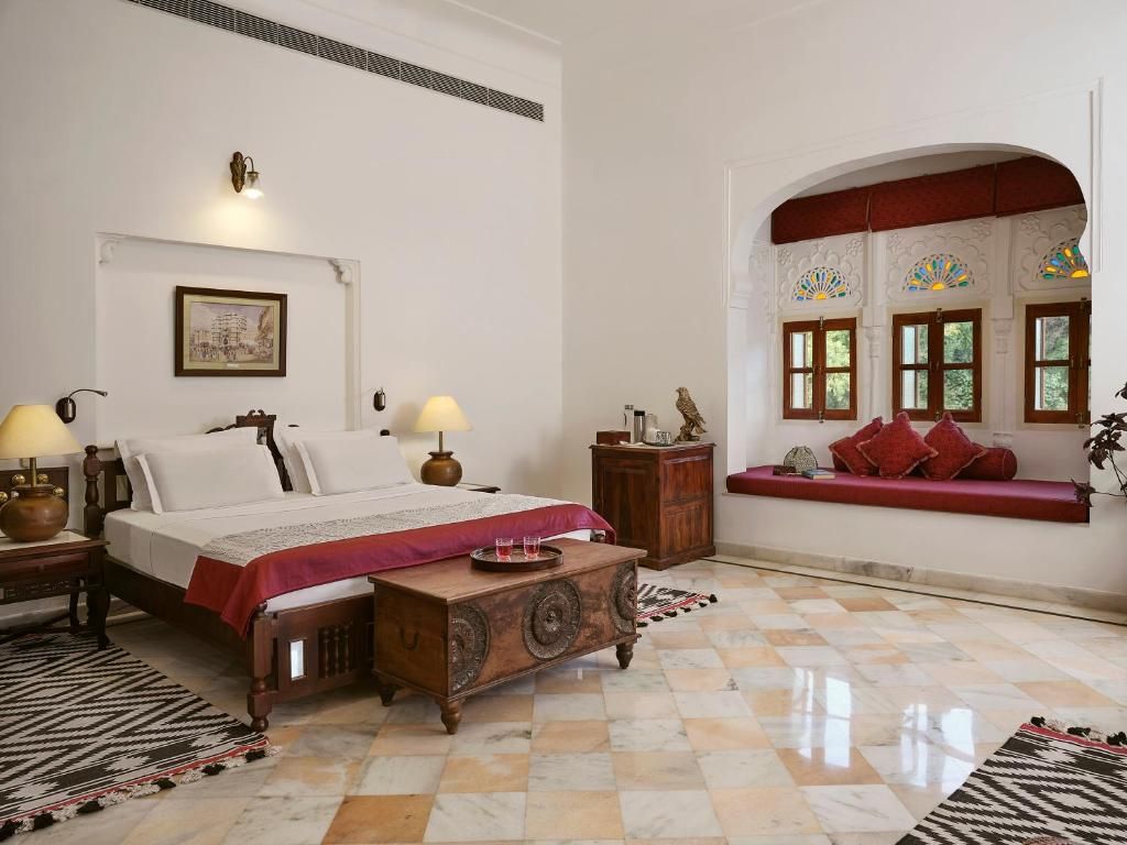 Um quarto espaçoso com uma cama grande, duas mesas de cabeceira, uma mesinha de madeira, um banco com almofadas vermelhas e uma janela decorativa com vitrais.