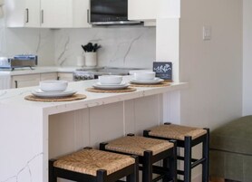 Uma cozinha com bancada branca, três banquetas com assentos trançados e um micro-ondas.
