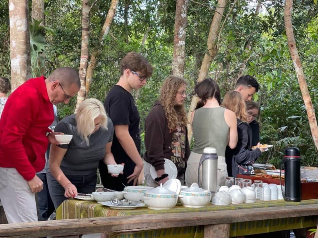 Um grupo de pessoas reunido em torno de uma mesa com diversos pratos e recipientes, provavelmente em um evento ou encontro ao ar livre.