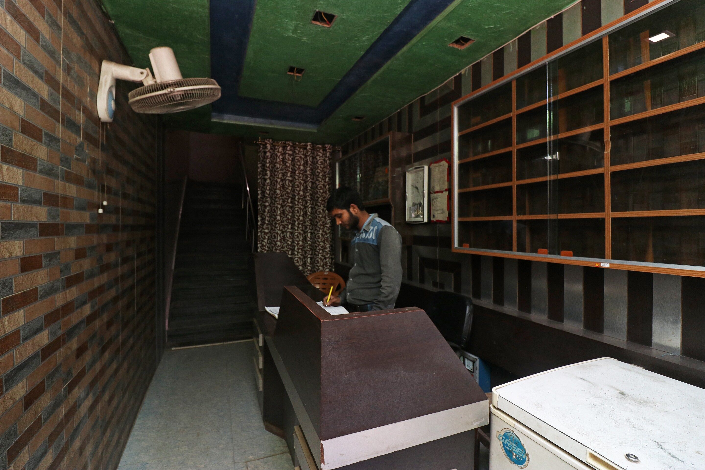 Balcão de recepção com uma pessoa sentada atrás, um ventilador fixado na parede e uma janela com persianas.